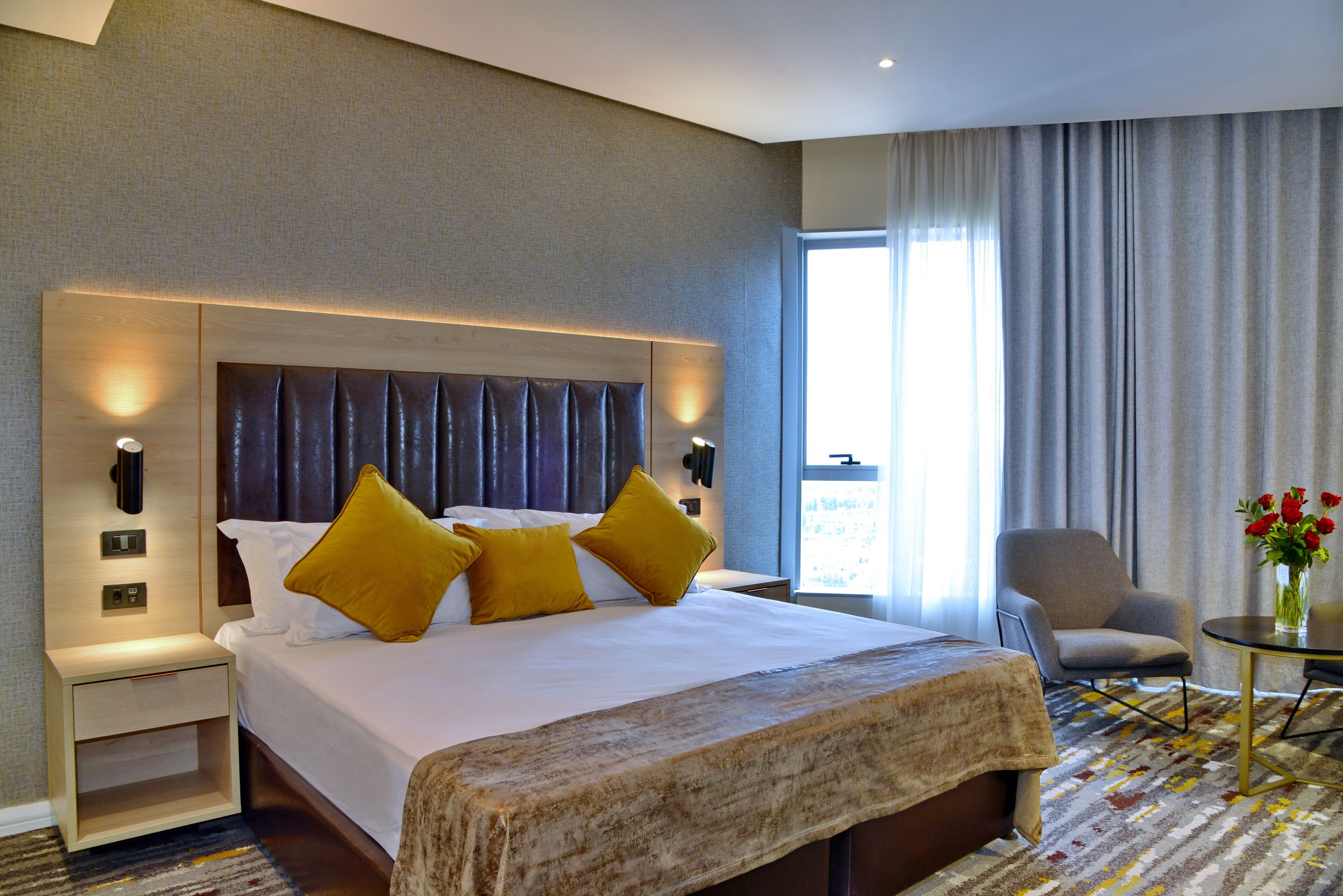 Quarto de hotel com uma cama grande, duas mesas de cabeceira, uma cadeira, uma mesinha com um vaso de flores e uma janela ampla com cortinas.