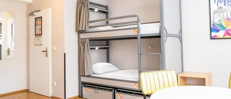 Uma cama de beliche com gavetas para guardar objetos, uma mesinha e uma cadeira num quarto com piso de madeira e uma porta.