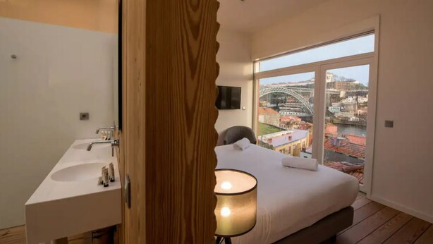 Quarto moderno com cama grande, porta de madeira, banheiro com pia e vista para a cidade através de amplas janelas.