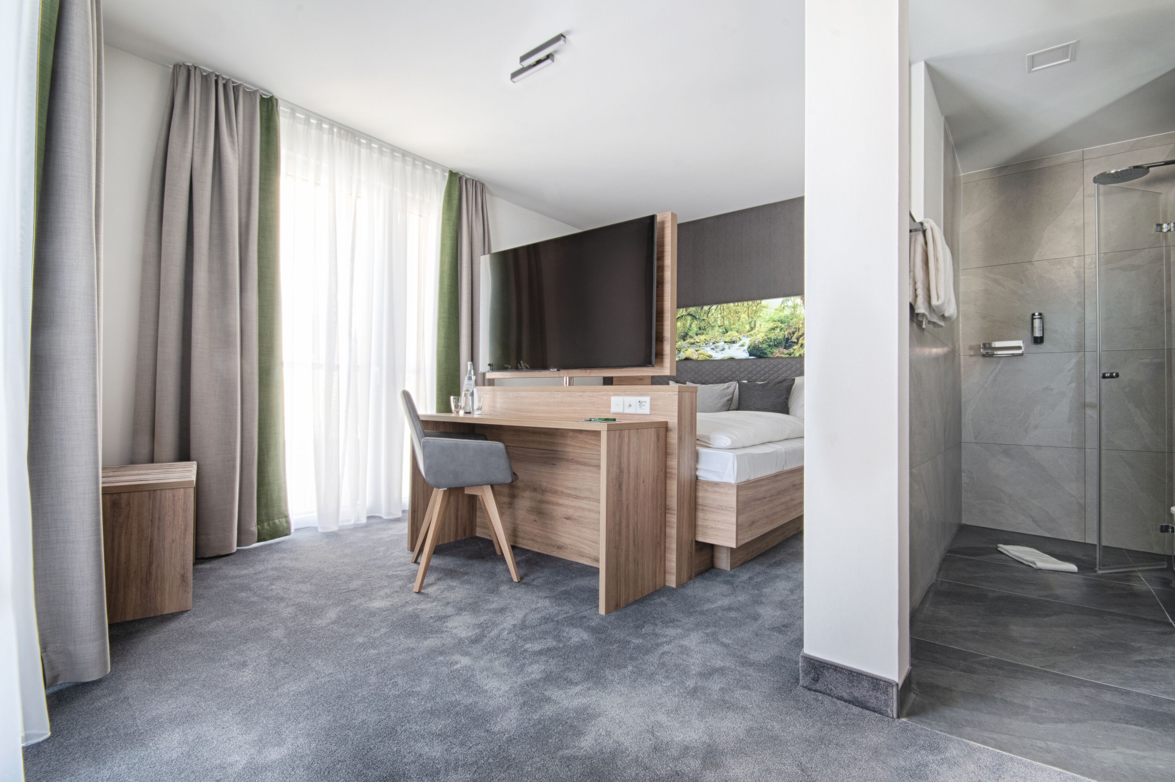 Quarto de hotel moderno com escrivaninha de madeira, cama, chuveiro e vista para o verde.