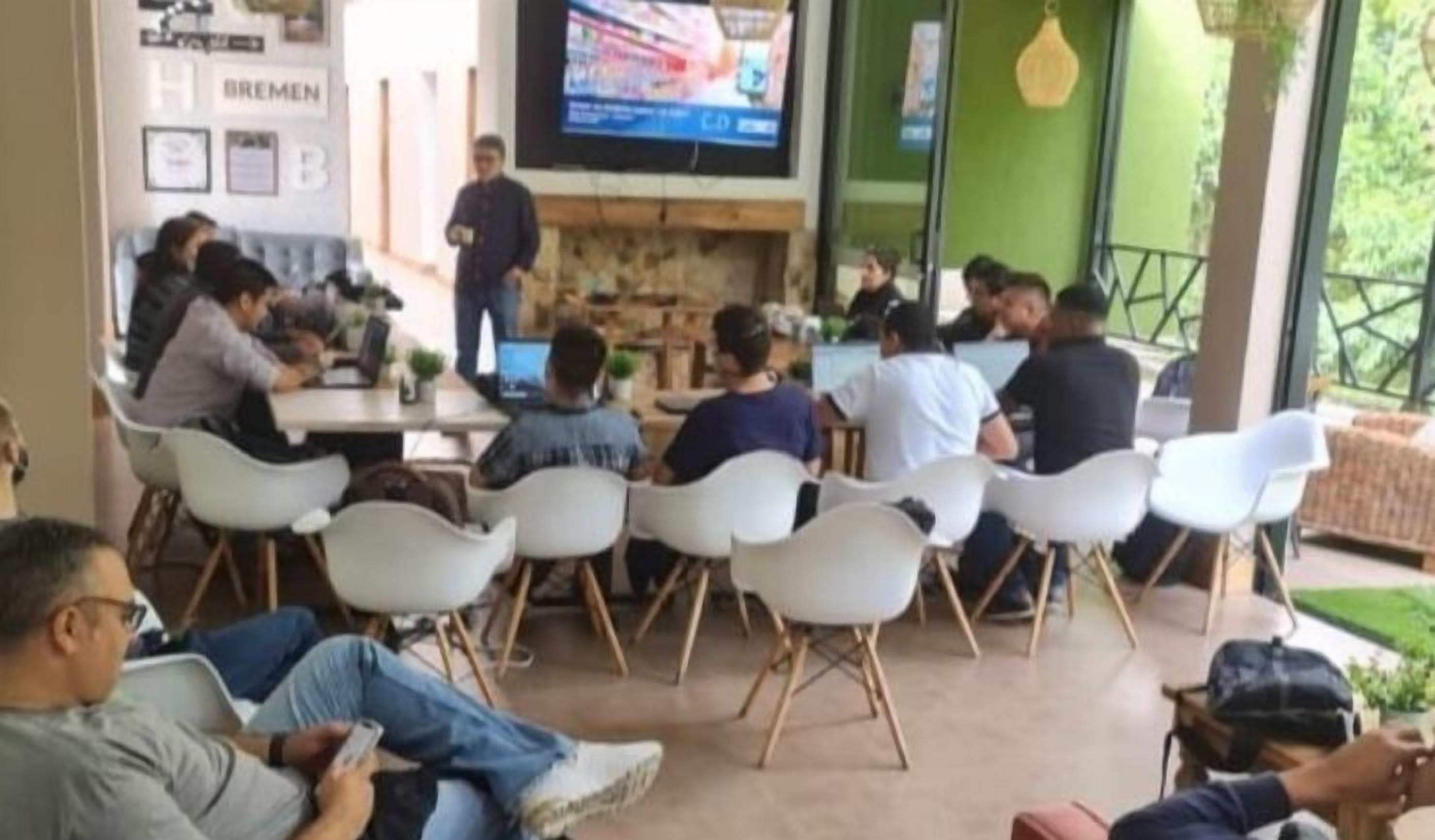Um grupo de pessoas sentadas em mesas numa sala com uma tela de televisão exibindo um canal de notícias.