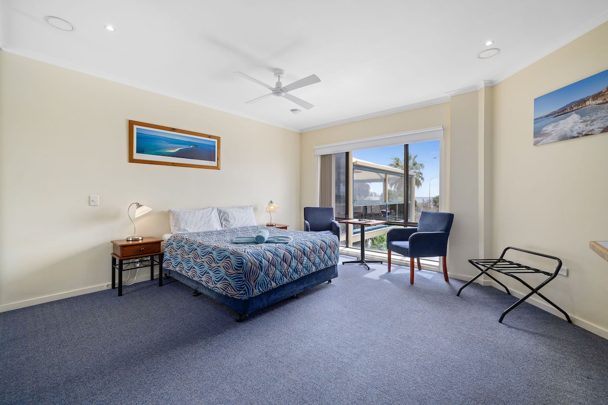 Quarto de hotel com cama, escrivaninha, cadeira, ventilador de teto e uma grande janela com vista.