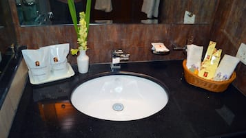 Casa de banho de hotel com lavatório branco, espelho e produtos de higiene numa cesta.