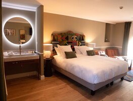 Quarto de hotel com uma cama grande, um espelho redondo, uma mesinha de cabeceira com abajur e um painel de cabeceira colorido.