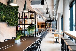 Um café moderno com um balcão comprido, cadeiras pretas e uma parede verde com plantas.