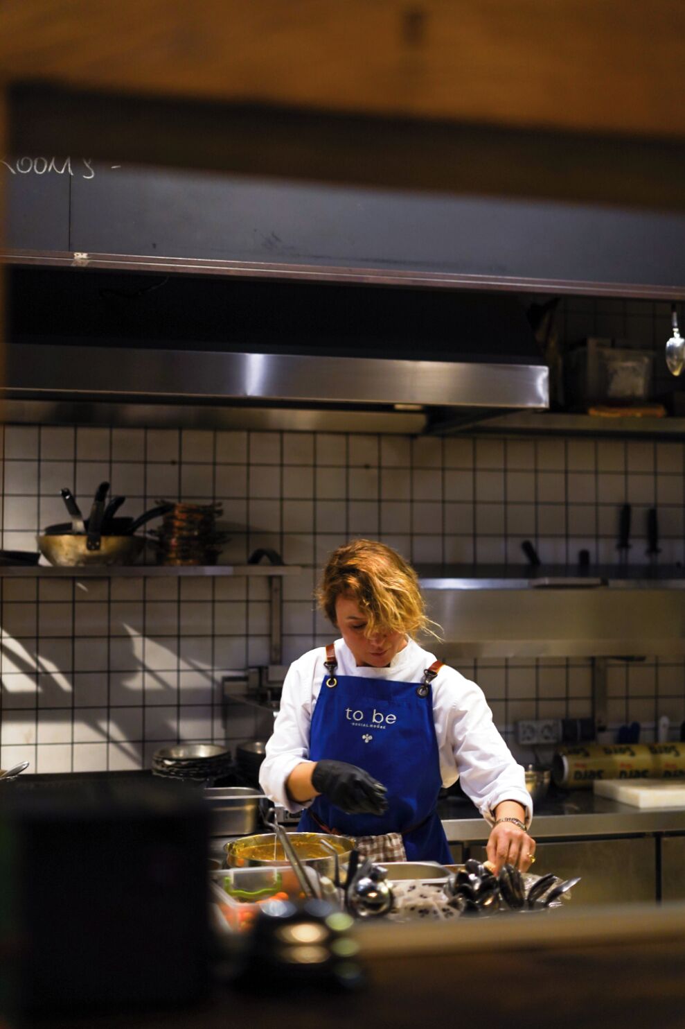 Uma pessoa em uma cozinha com avental azul, eletrodomésticos de aço inoxidável e painel de azulejos.