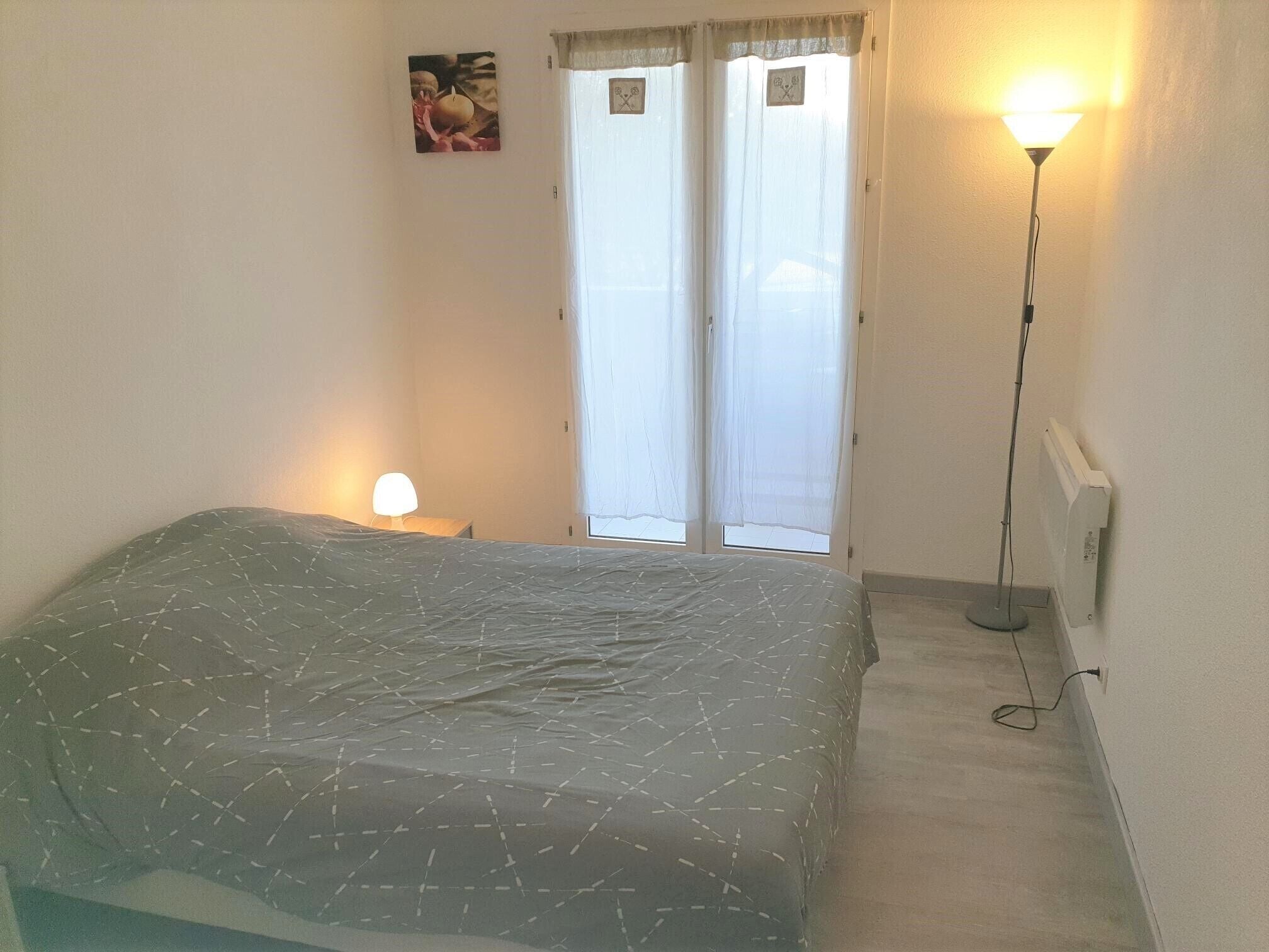 Quarto com cama, mesa de cabeceira, abajur e janela com cortinas.
