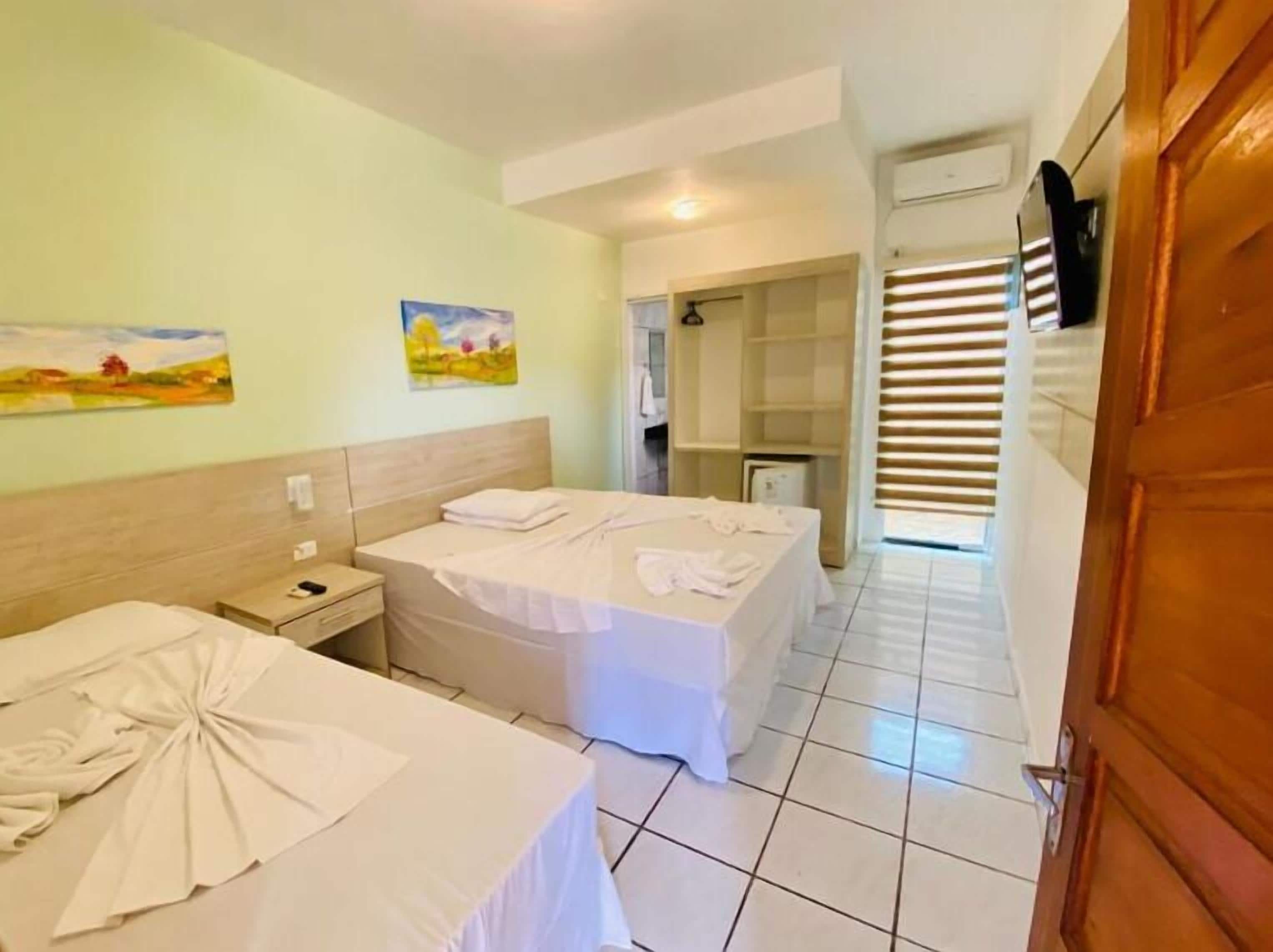Quarto de hotel com duas camas, uma mesa de cabeceira, um quadro na parede e um banheiro visível através de uma porta de vidro.