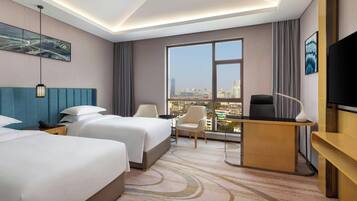 Quarto de hotel com duas camas, uma escrivaninha, uma cadeira, televisão e uma janela grande com vista para a cidade.