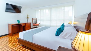 Quarto de hotel com uma cama, escrivaninha, cadeira, televisão e janela com cortinas.