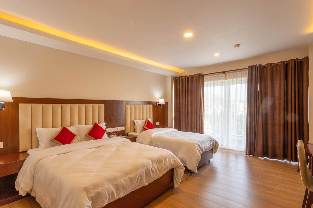 Quarto de hotel com duas camas, cabeceira de madeira, paredes bege e uma janela grande com cortinas.
