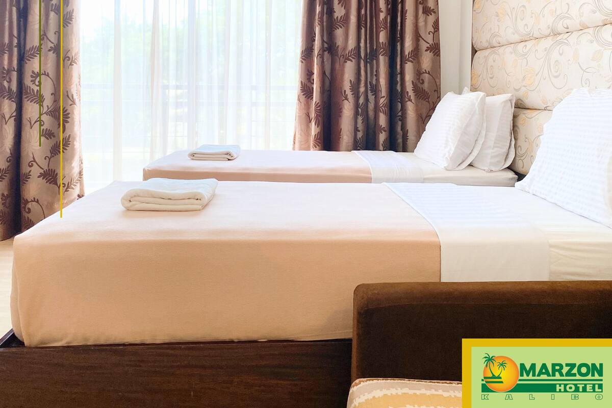 Quarto de hotel com duas camas, cabeceira com padrão floral e janela com cortinas translúcidas.