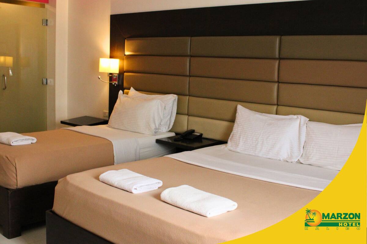 Quarto de hotel com duas camas, cada uma com lençóis e almofadas brancas, uma mesinha de cabeceira com abajur e um guarda-roupa.