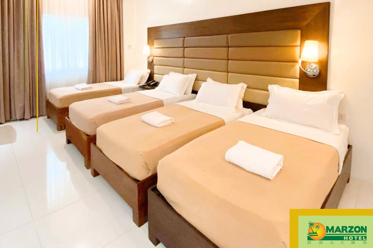 Quarto de hotel com quatro camas dispostas em linha, uma cabeceira grande e uma janela com cortinas.