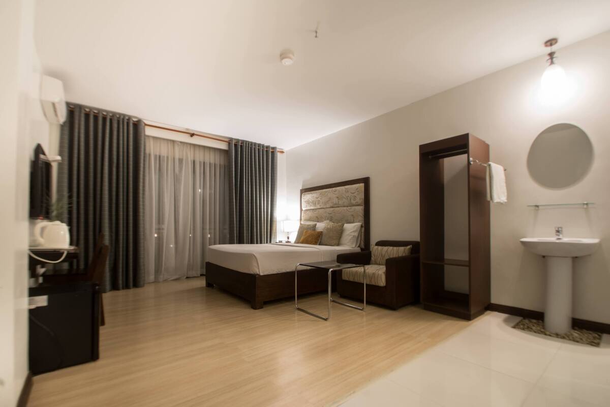 Quarto de hotel moderno com cama, poltrona, escrivaninha, espelho, pia e janela com cortinas.
