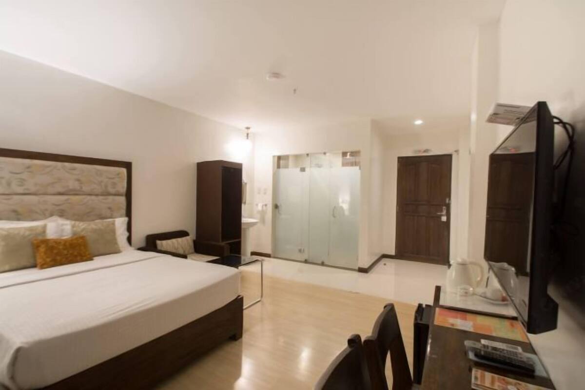 Quarto de hotel com uma cama, uma escrivaninha com cadeira, televisão, guarda-roupa e banheiro com porta de vidro.