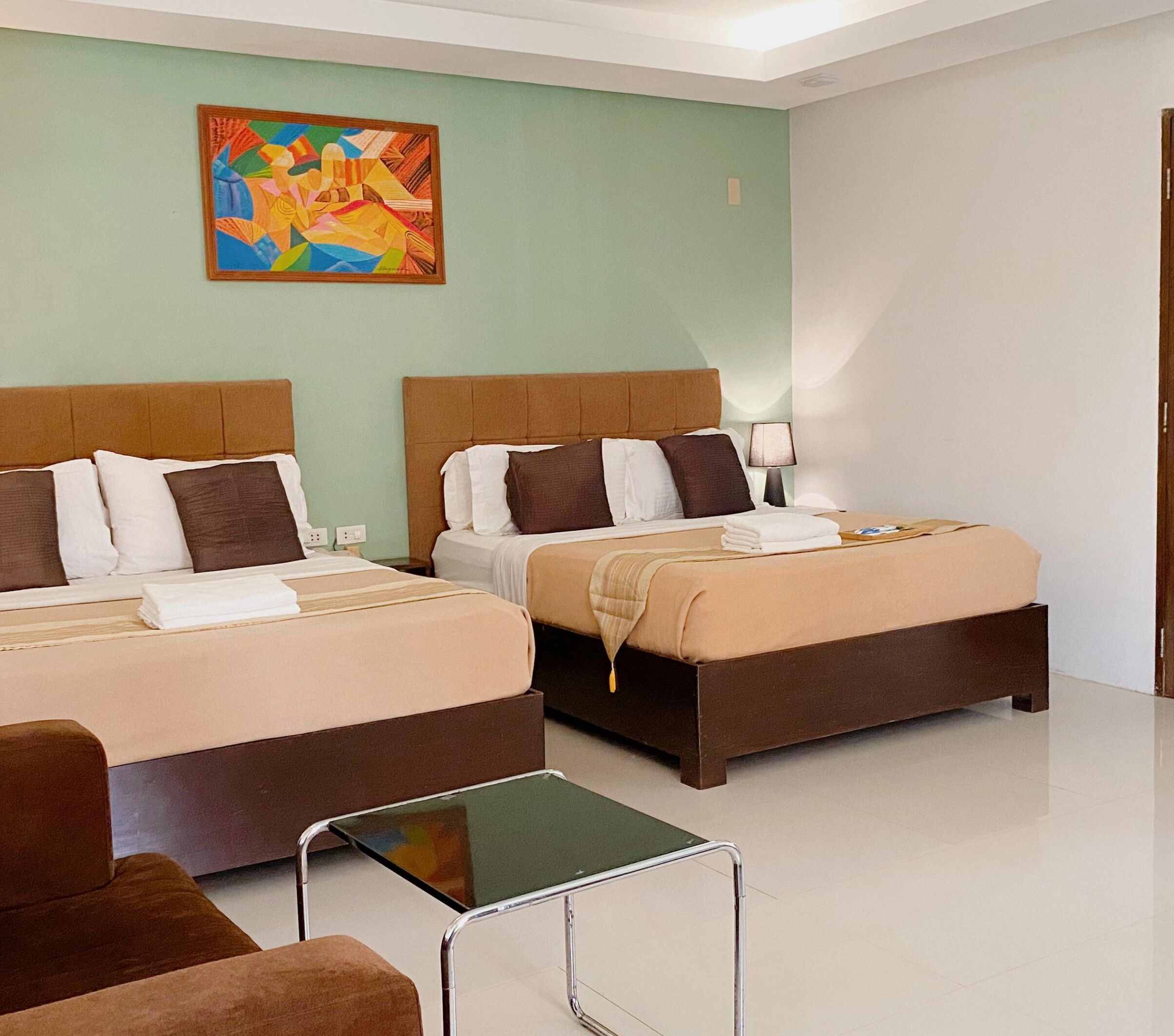 Quarto de hotel com duas camas, uma mesinha e um quadro abstrato colorido na parede.