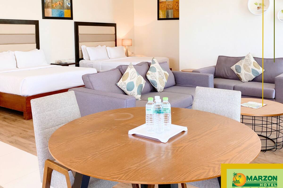 Quarto de hotel com uma cama, dois sofás, uma mesa redonda de madeira com garrafas de água e duas luminárias fixadas na parede.