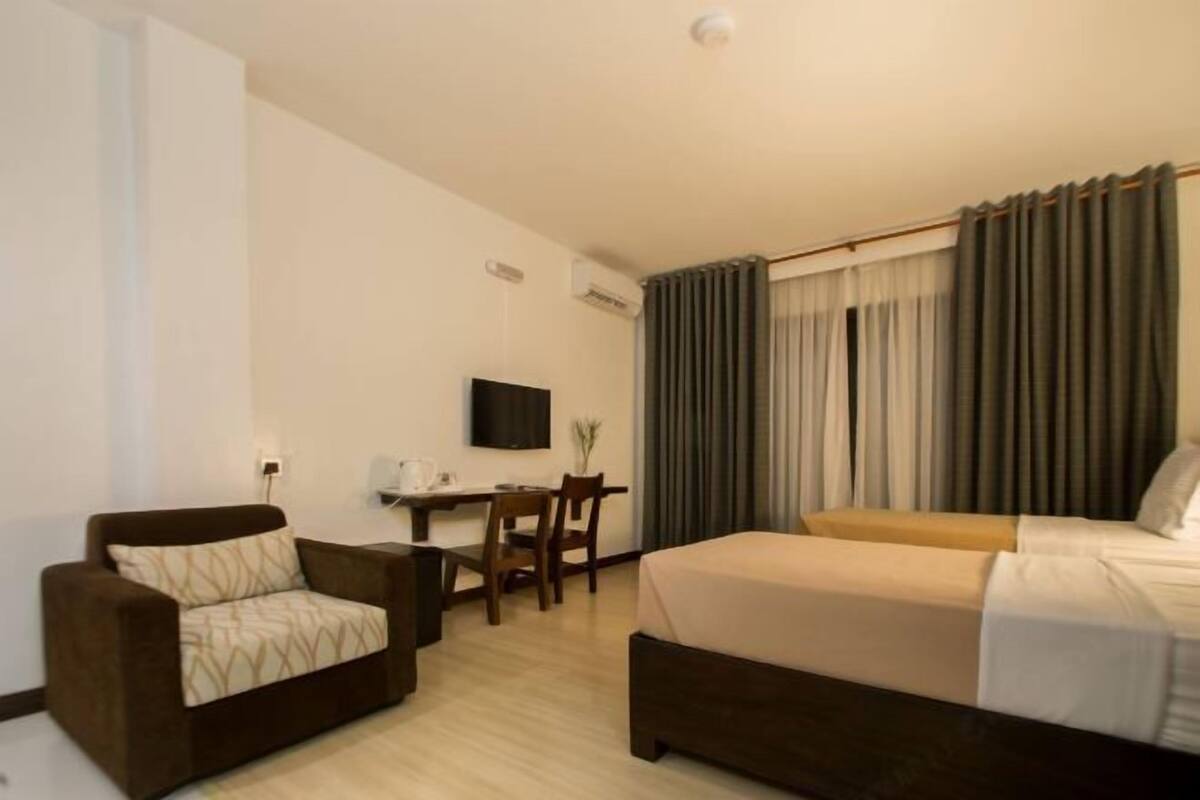 Quarto de hotel com cama, sofá, escrivaninha com cadeira, televisão e cortinas.