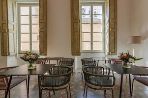Uma sala de jantar com mesas e cadeiras de vime, grandes janelas com persianas e um vaso com flores sobre a mesa.