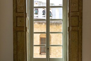 Visão através de uma janela de madeira, revelando um edifício de pedra com várias janelas e um campanário.