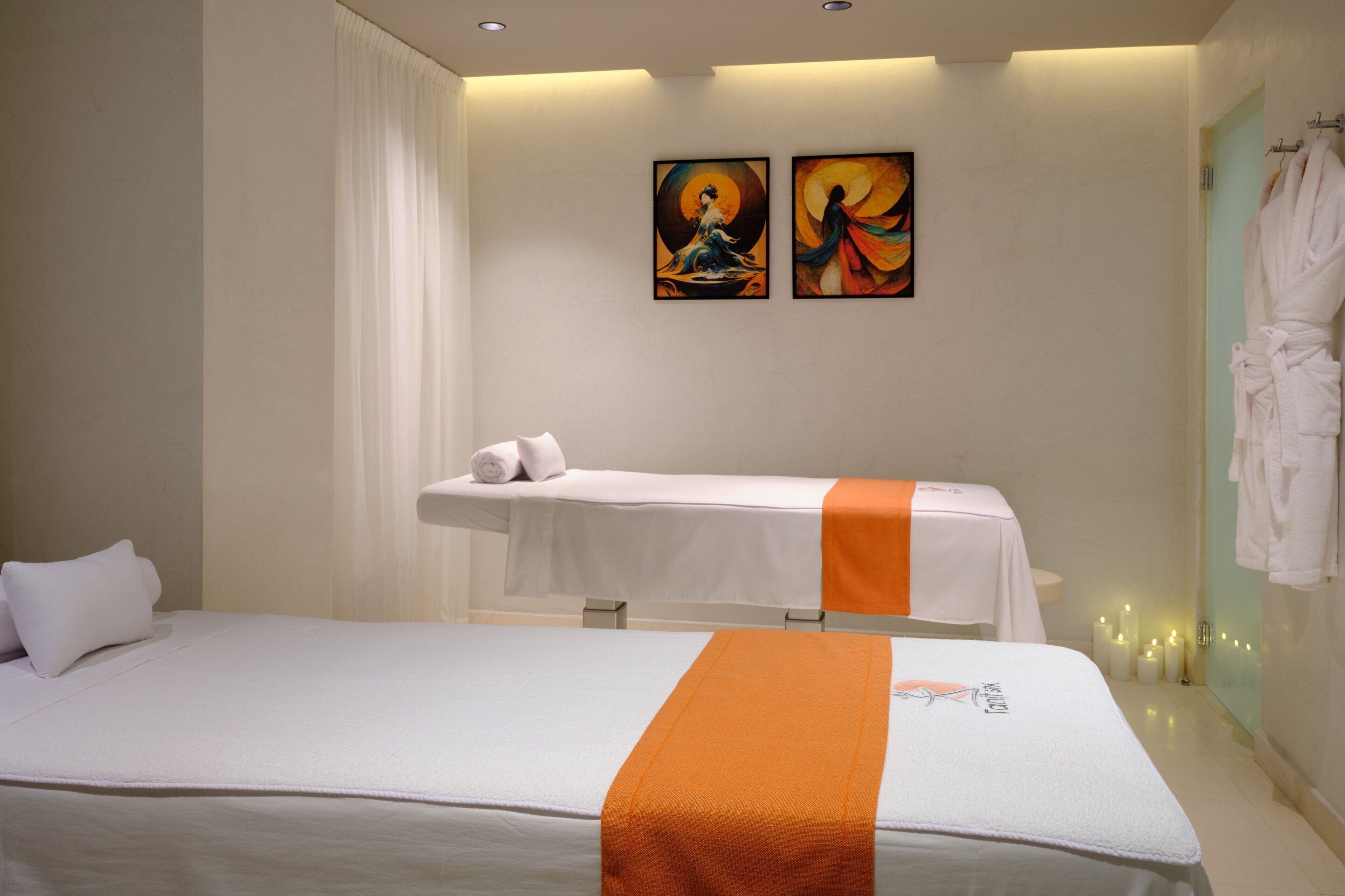 Quarto de hotel com duas camas, mesa de massagem, obras de arte na parede e um roupão pendurado na porta.