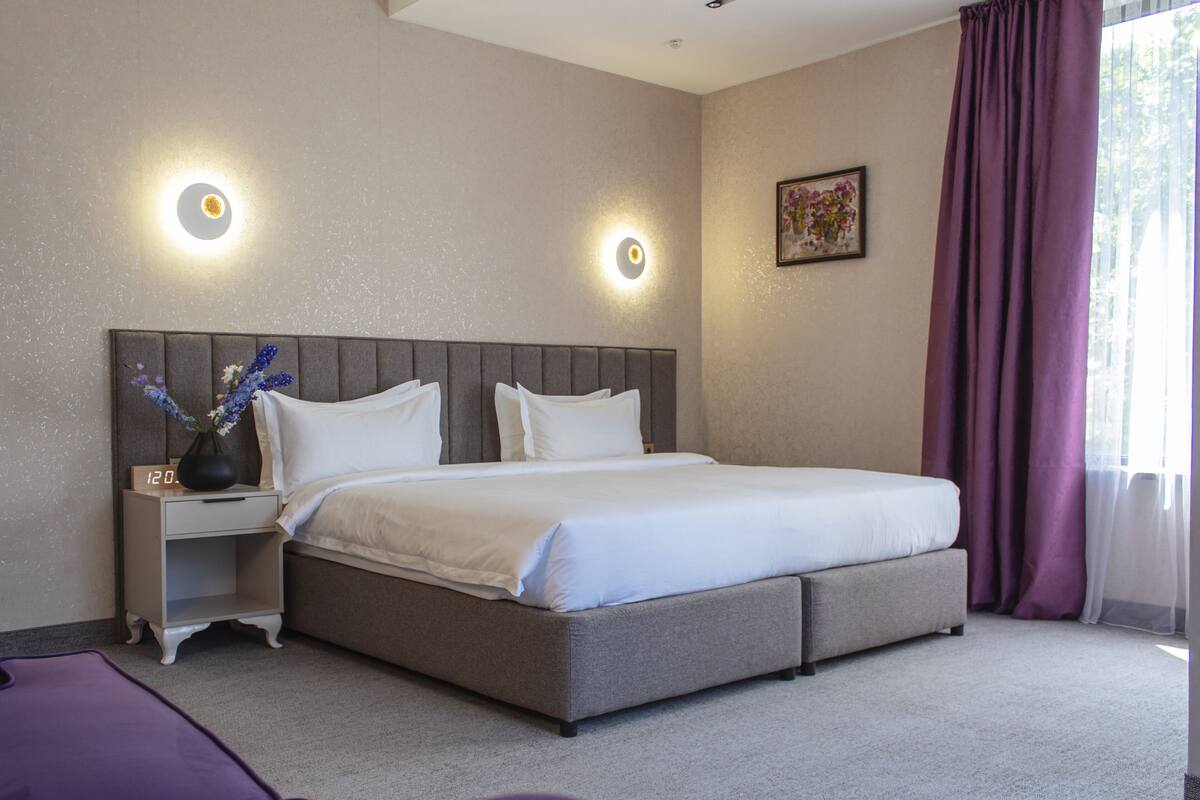 Quarto de hotel moderno com uma cama grande, duas mesas de cabeceira, um criado-mudo, um vaso com flores e um quadro na parede.