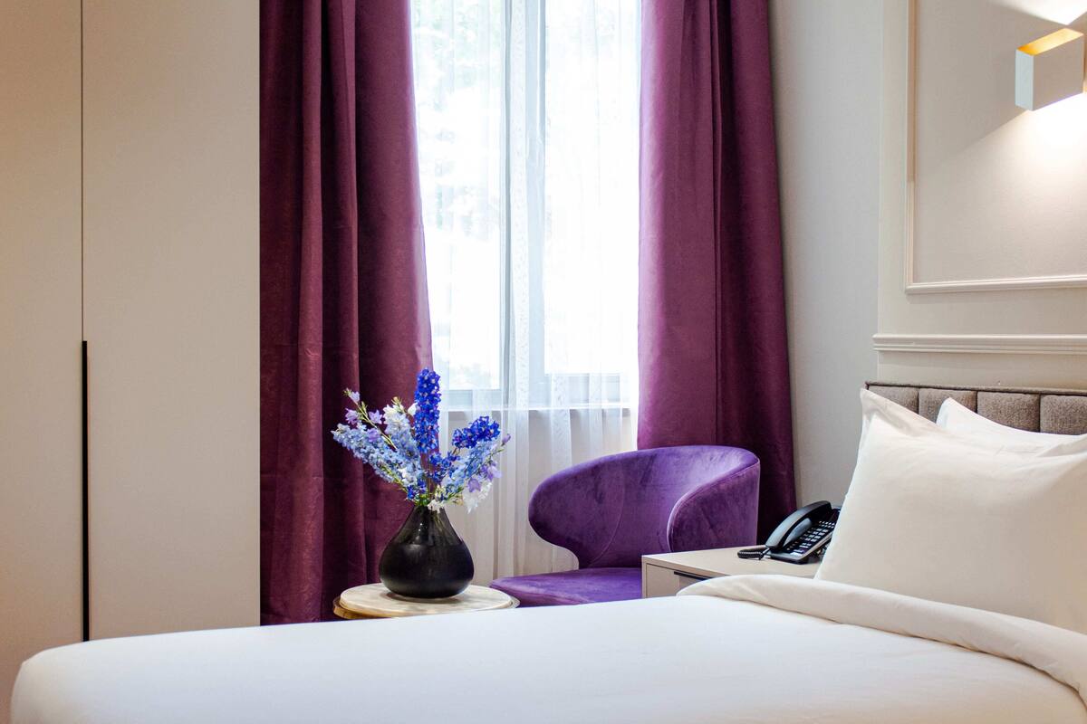 Quarto de hotel com cama, mesa de cabeceira, um vaso com flores, uma cadeira roxa e uma janela com cortinas.