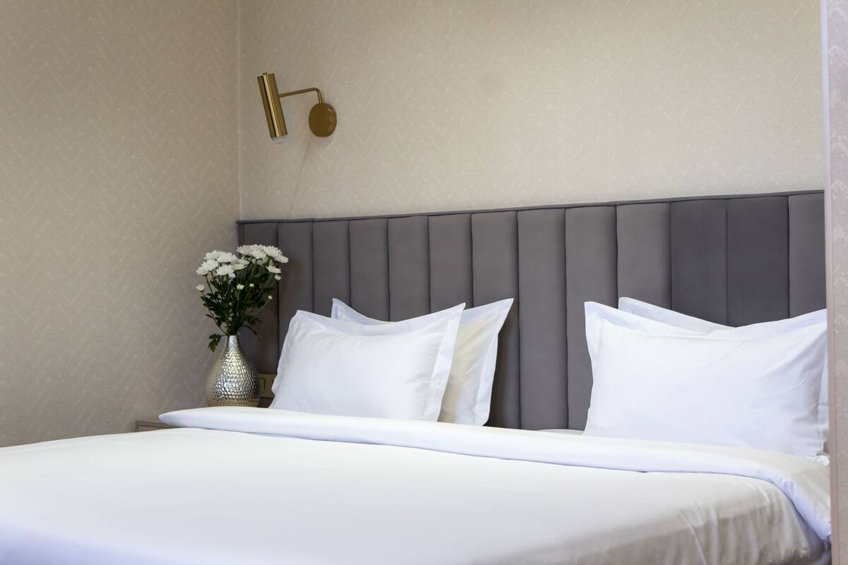 Cama bem arrumada com lençóis e almofadas brancas, um vaso com flores sobre uma mesa de cabeceira e um luminário fixado na parede.