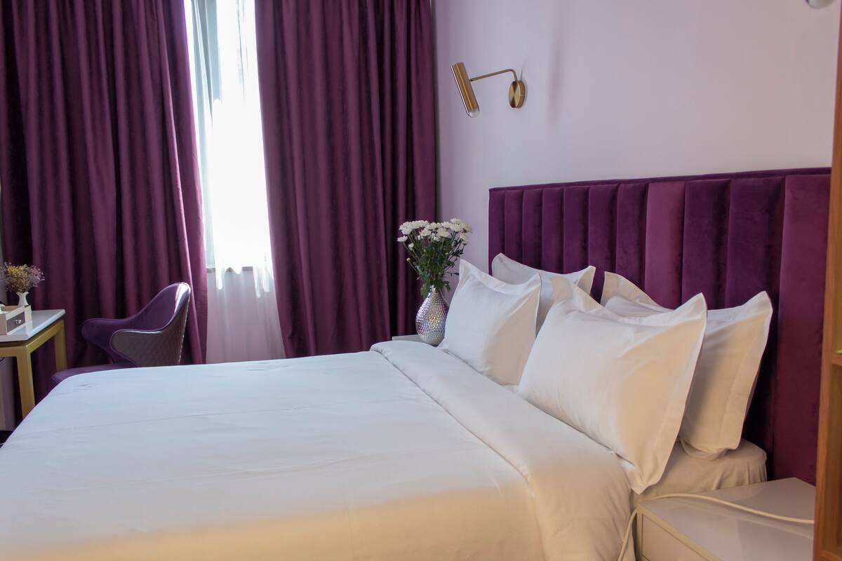 Quarto de hotel com uma cama grande, uma mesa de cabeceira, uma cadeira e um vaso com flores.