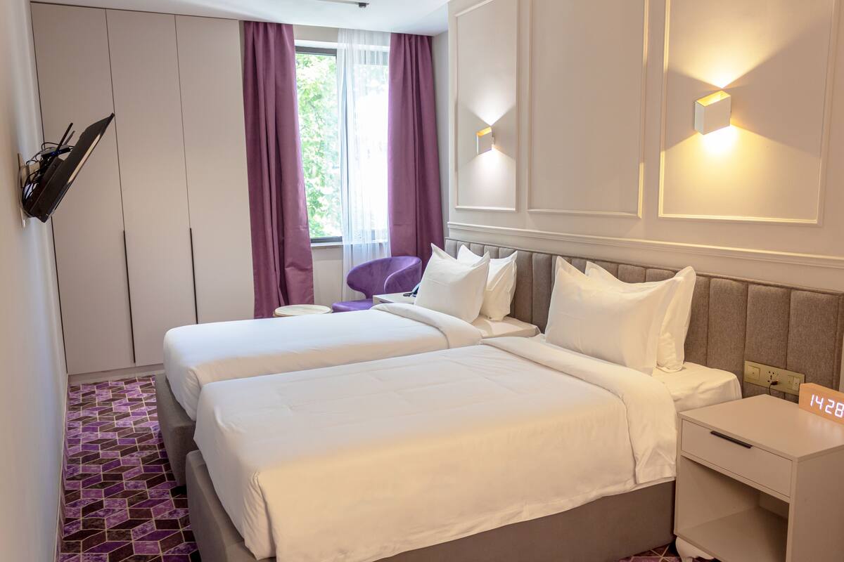 Quarto de hotel com duas camas, uma mesa de cabeceira, uma luminária na parede e uma janela com cortinas.