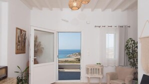 Quarto com uma grande porta de correr que dá para uma varanda com vista para o mar. Há um sofá branco, uma mesa pequena e um vaso de plantas.
