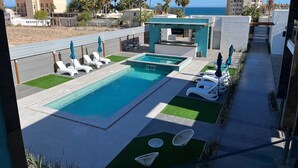 Uma área de piscina moderna ao ar livre, com espreguiçadeiras e uma piscina pequena, rodeada por palmeiras e edifícios.