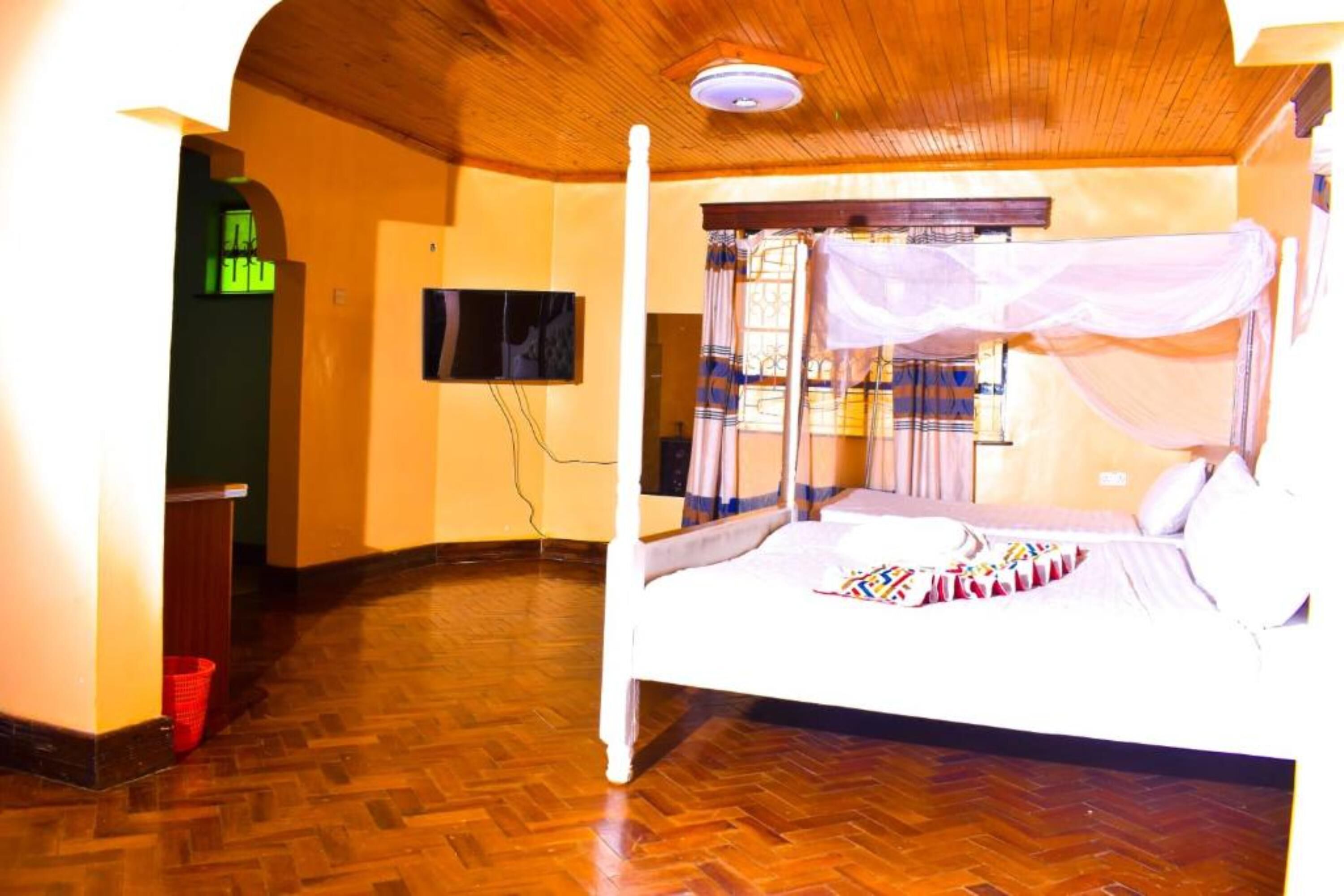 Quarto com cama de dossel, piso de madeira, televisão fixada na parede e janela com cortinas.
