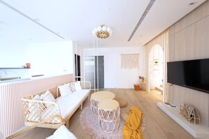 Uma sala de estar moderna com um sofá, duas mesas redondas, uma televisão de tela plana e uma cozinha ao fundo.