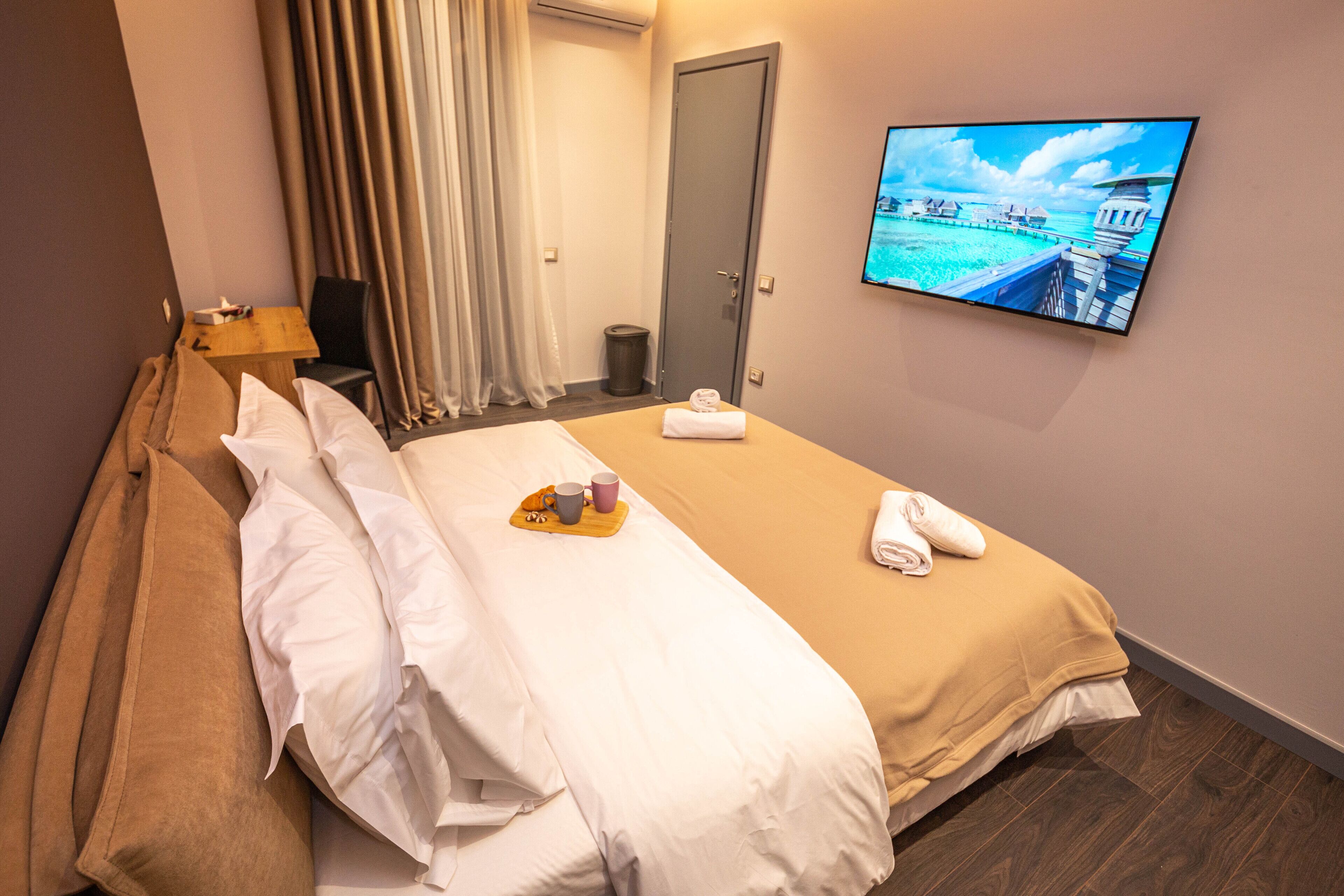Quarto de hotel com cama, televisão na parede, escrivaninha com cadeira e janela com cortinas.