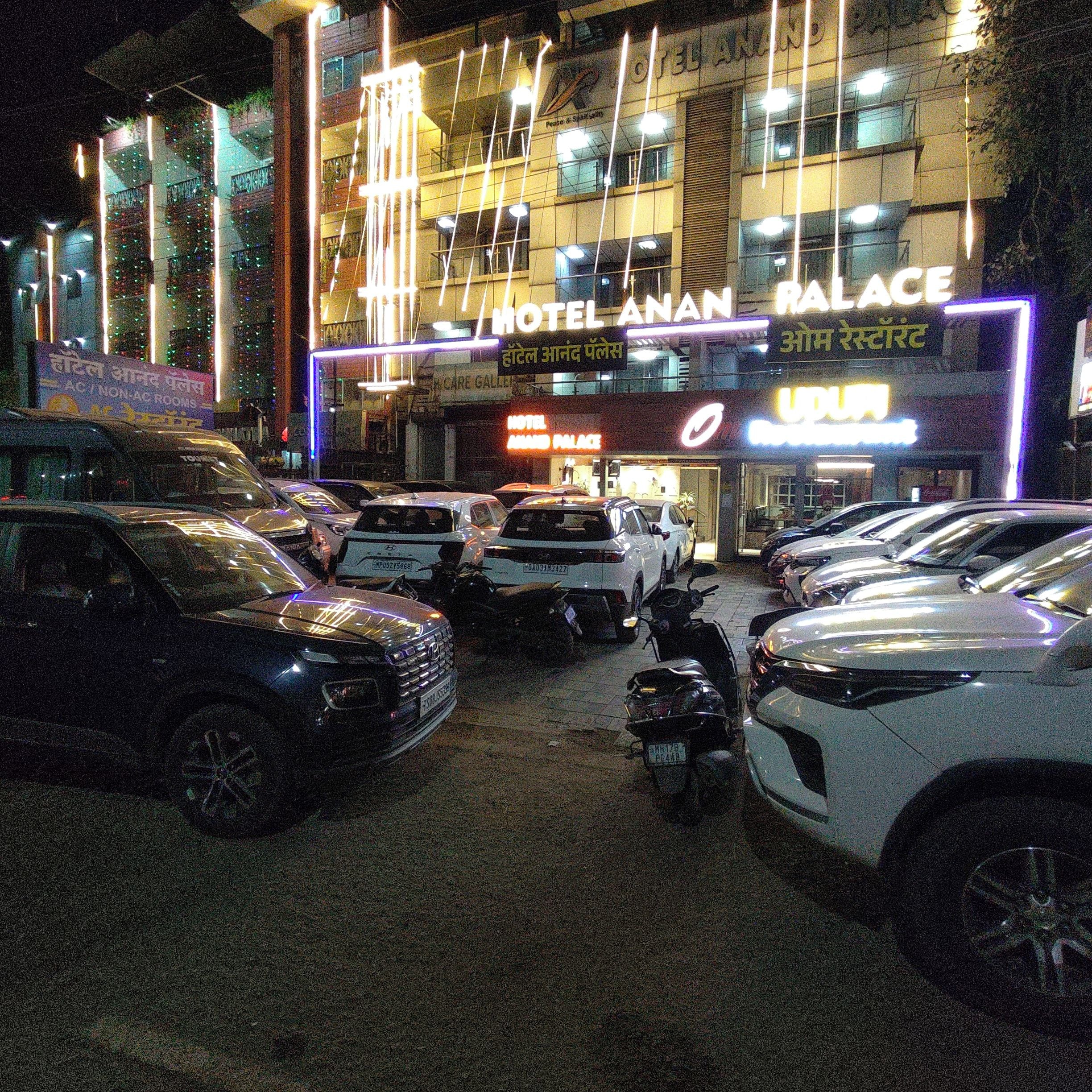 Um hotel com vários andares, um estacionamento com diversos carros e uma placa indicando 'Hotel Anan Palace'.