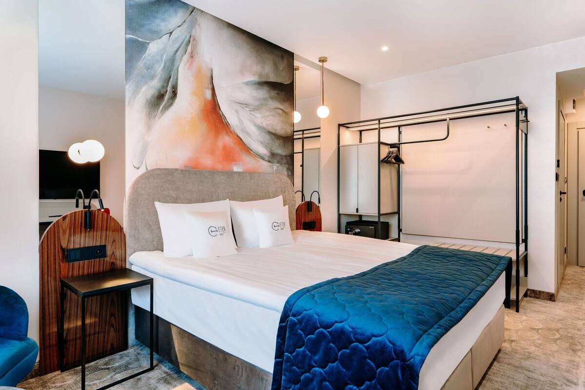 Quarto de hotel moderno com uma cama grande, uma mesinha de madeira, um guarda-roupa e um grande quadro abstrato na parede.