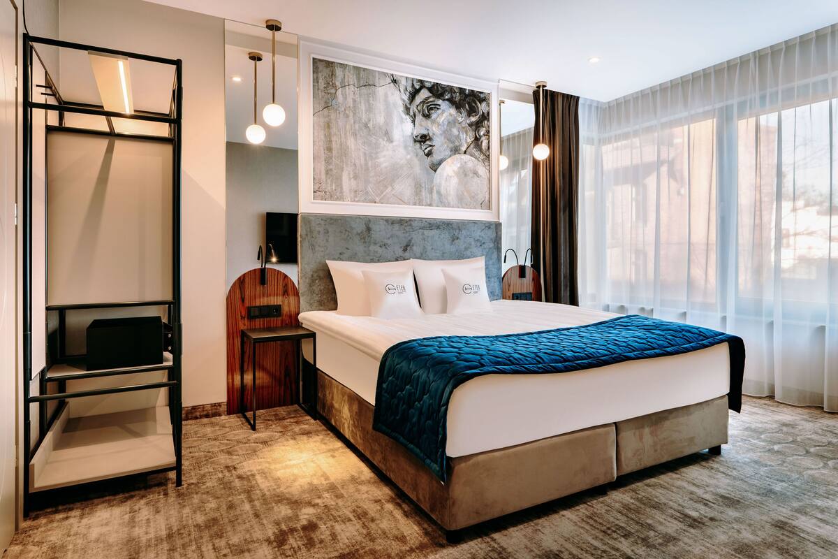 Quarto de hotel moderno com uma cama grande, um guarda-roupa, uma escrivaninha e uma janela ampla com cortinas.