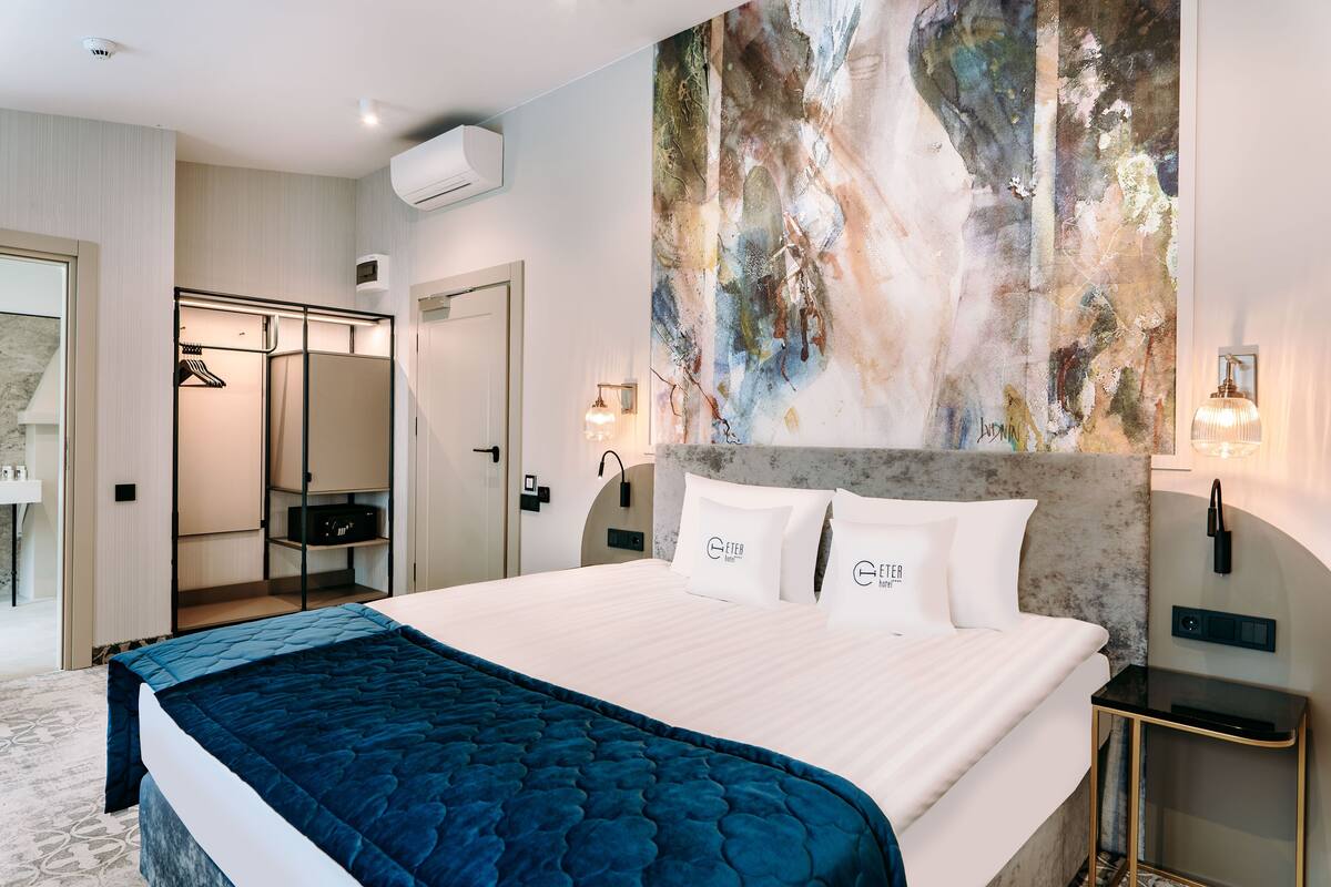 Quarto de hotel com uma cama grande, um guarda-roupa, um quadro na parede e uma mesa de cabeceira com abajur.