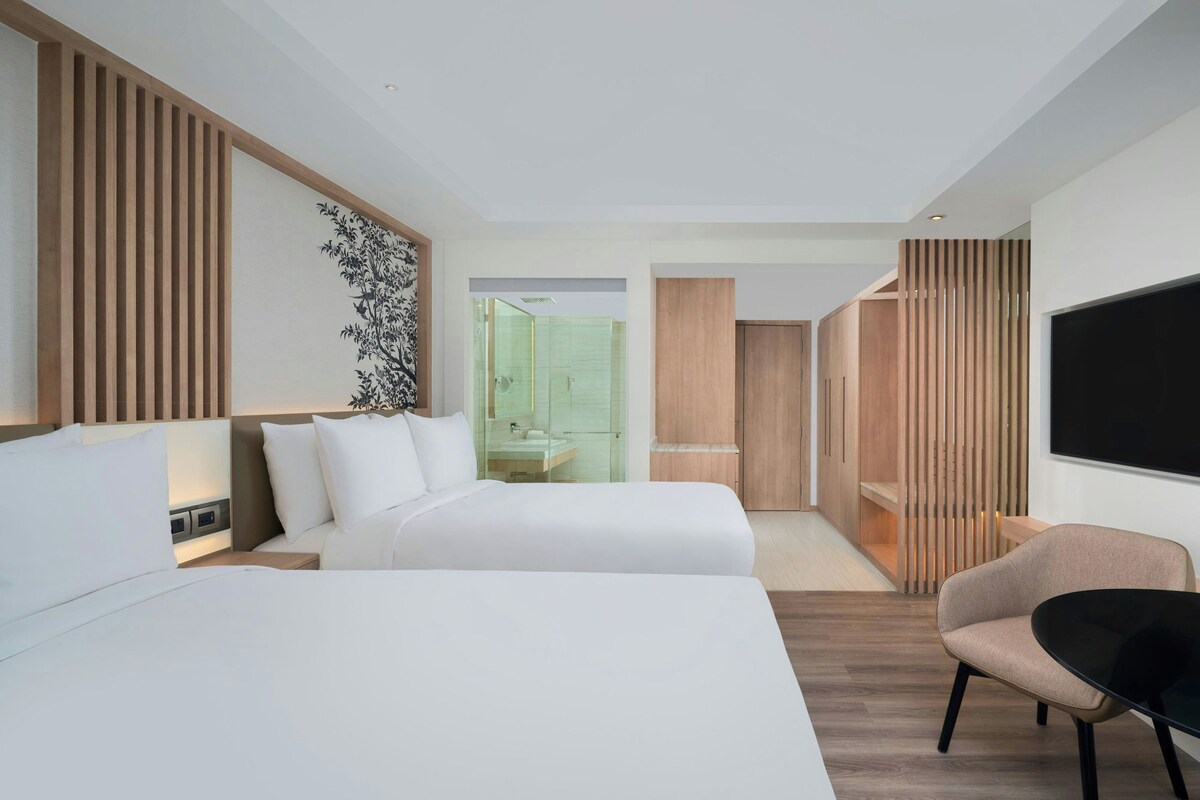 Quarto de hotel moderno com duas camas, uma mesa, uma cadeira, uma TV e um banheiro visível através de um painel de vidro.
