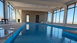 Piscina interior com água azul cristalina, grandes janelas e um banco.