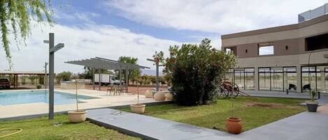 Uma área de piscina com um pérgola, mobiliário de exterior e um edifício com grandes janelas.