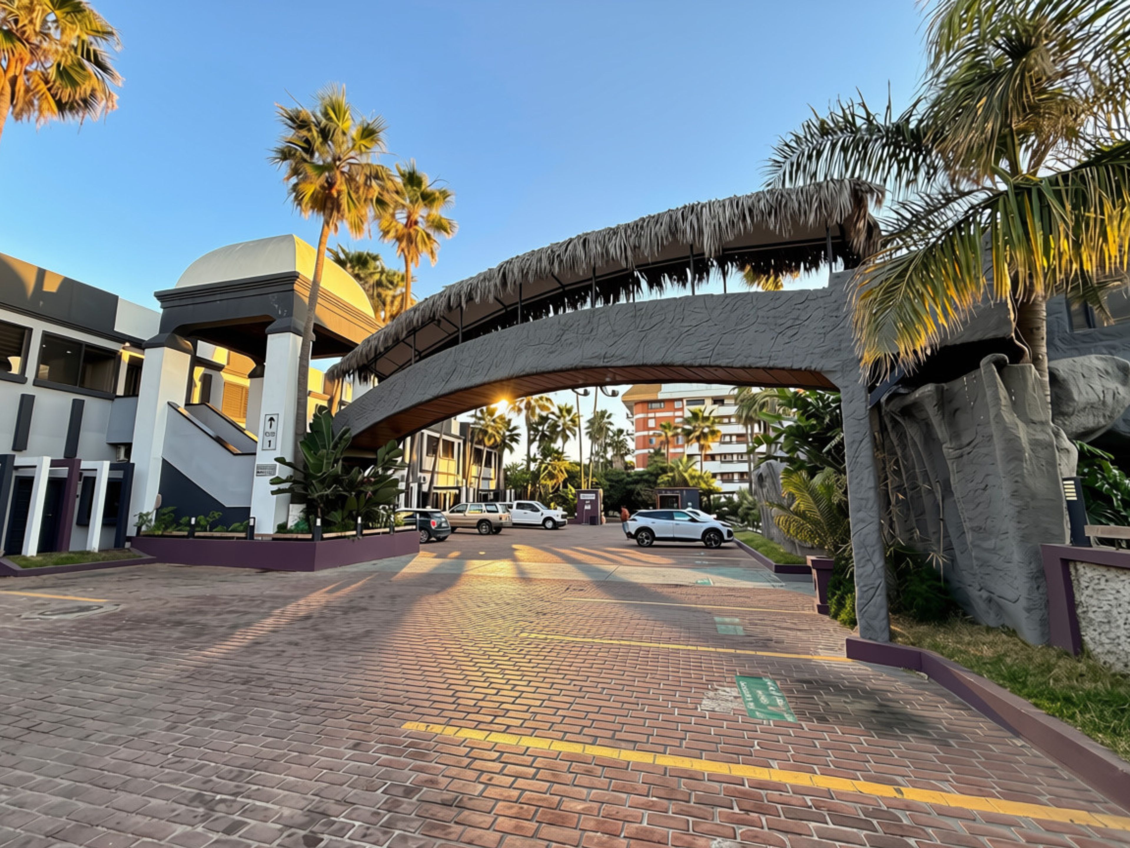 Uma entrada pavimentada com um arco de palha, palmeiras e um edifício de vários andares com design moderno.
