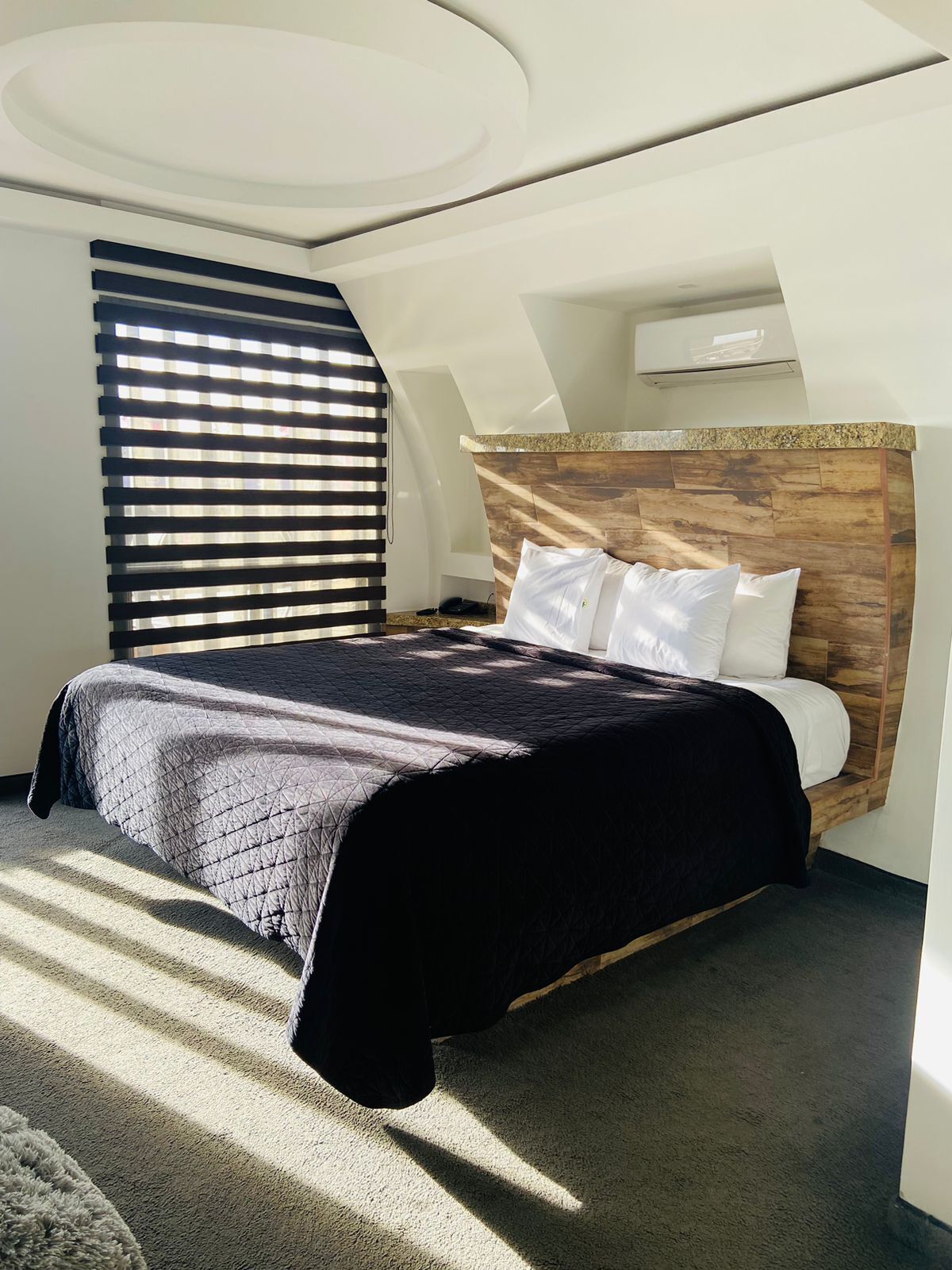Quarto moderno com teto curvo, cama com cobertura acolchoada escura, cabeceira de madeira e uma janela grande com persianas.