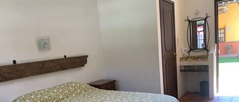 Quarto com cama, cabeceira de madeira, mesa de cabeceira pequena, porta e banheiro com pia e espelho.