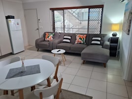Uma sala de estar moderna com um sofá cinza, uma mesa de jantar com cadeiras, uma geladeira branca e uma janela com persianas.