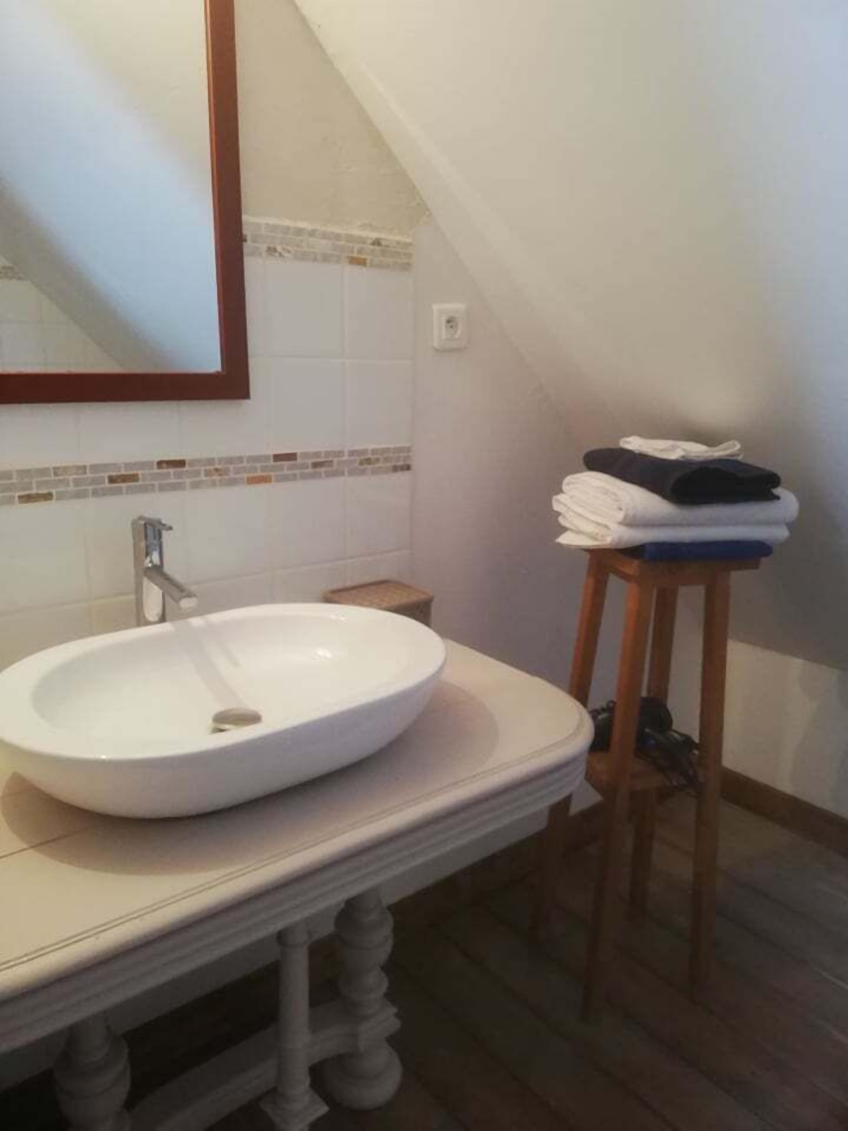 Uma casa de banho com lavatório branco, um espelho e um pequeno banco de madeira com toalhas dobradas.