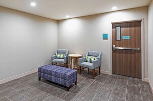 Quarto de hotel com porta de madeira, duas poltronas, uma mesa redonda pequena e um banquinho otomano azul estampado.
