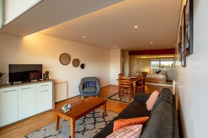Uma sala de estar moderna com sofá, mesa de centro e TV sobre um móvel. Há também uma área de jantar com mesa e cadeiras.